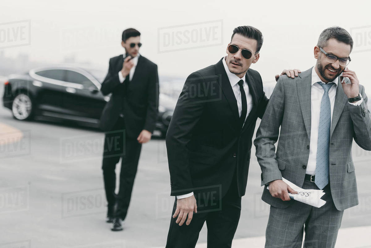 Happy businessman walking with bodyguards and talking by smartphone ...