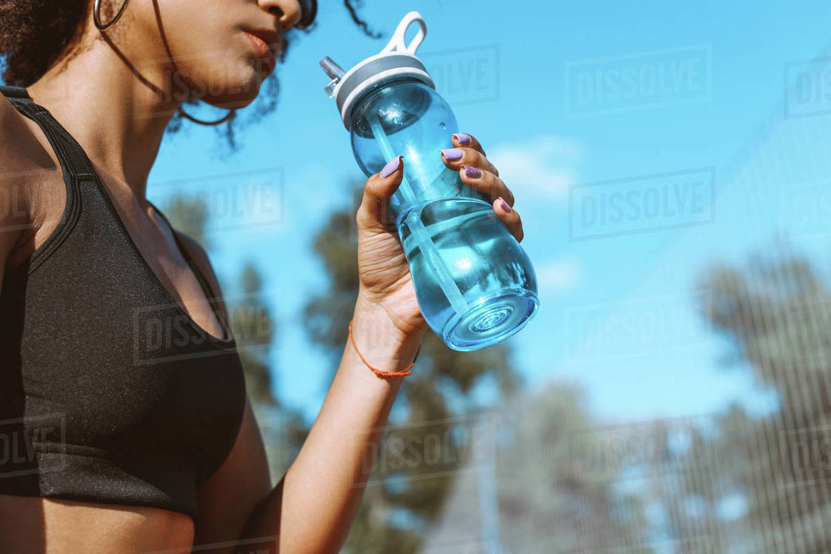 Cropped shot of woman in sports bra drinking from blue water bottle ...