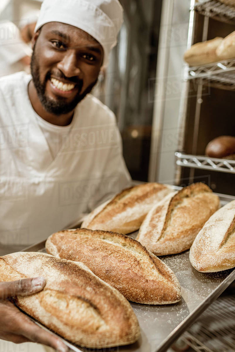 Smiling handsome african american baker with tray of fresh loaves of ...