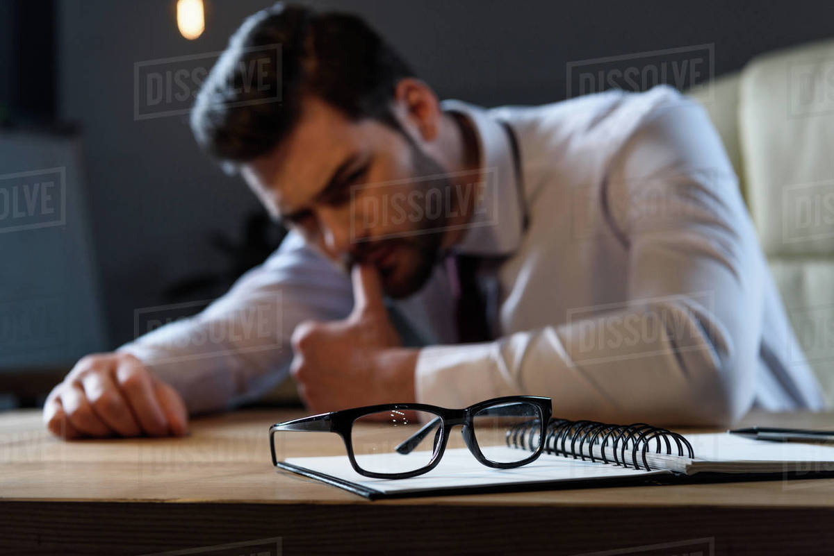 Tired pensive businessman leaning on table with glasses on foreground ...