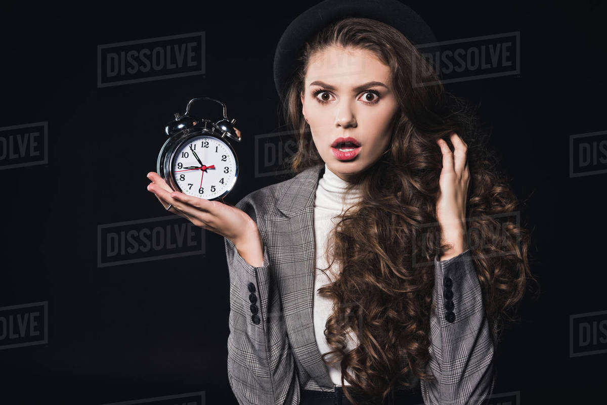 Shocked young woman holding clock and looking at camera isolated on ...