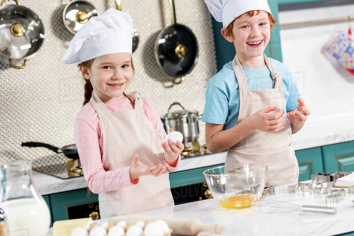 Adorable happy children in chef hats and aprons smiling at camera while ...
