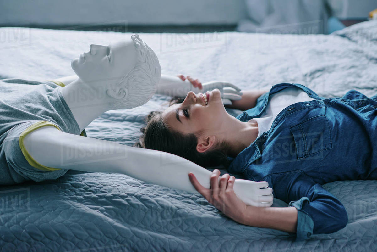 Side view of smiling woman and mannequin lying on bed, loneliness ...