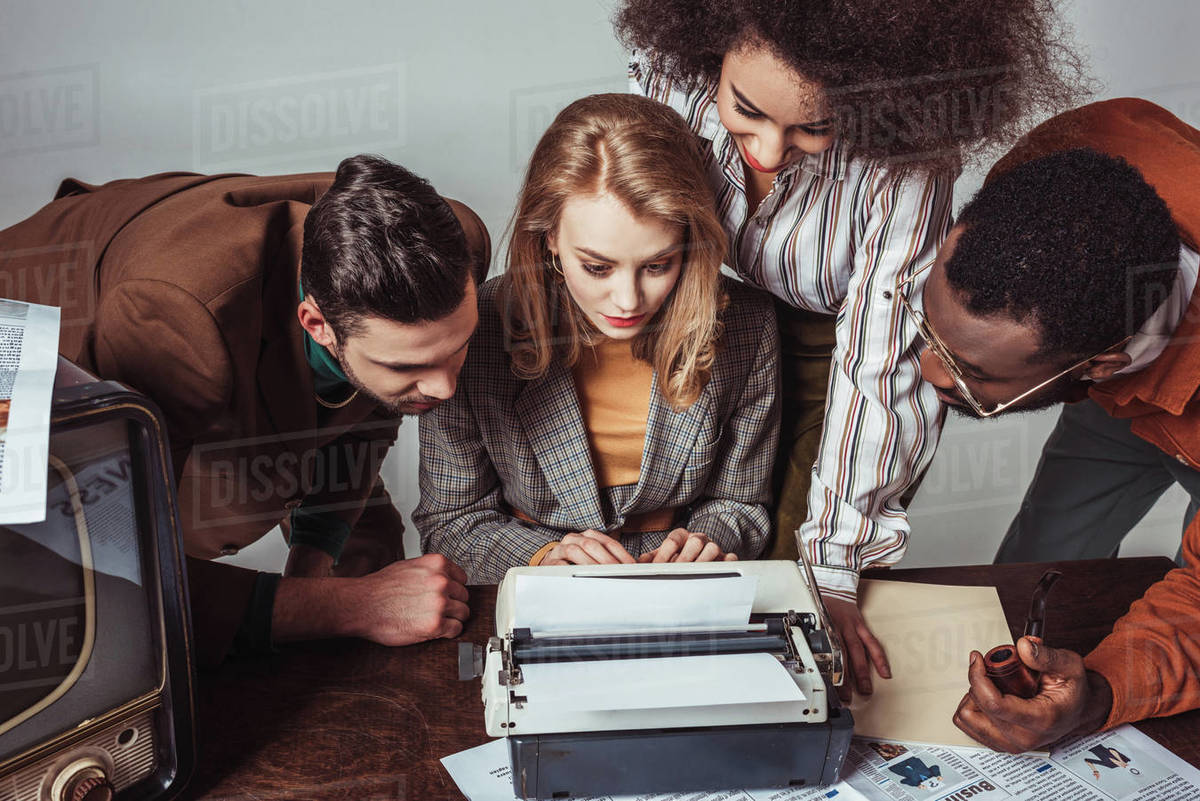 Multicultural retro styled journalists looking at text at typewriter ...