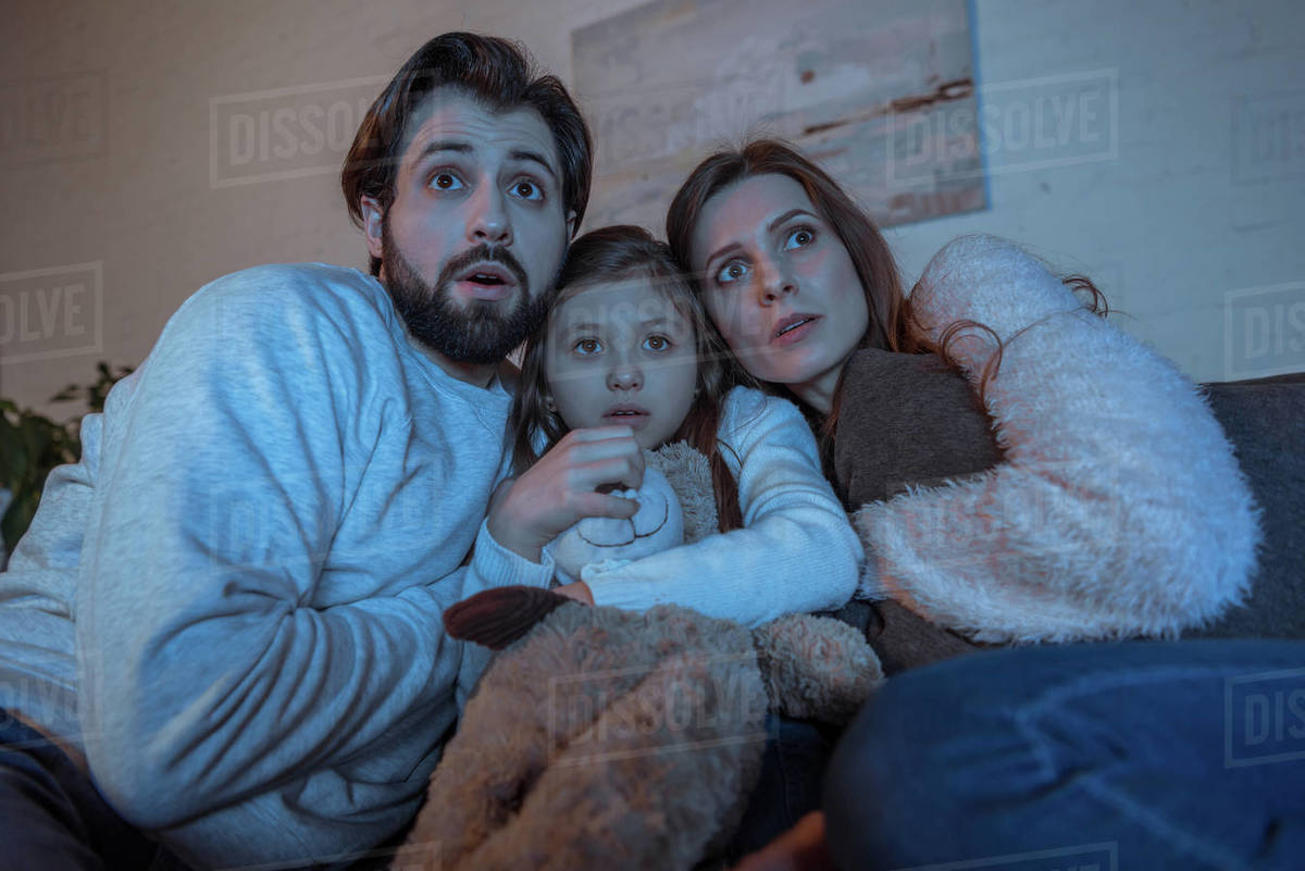 Scared parents and daughter watching horror movie in evening - Royalty-free Stock Photo | Dissolve
