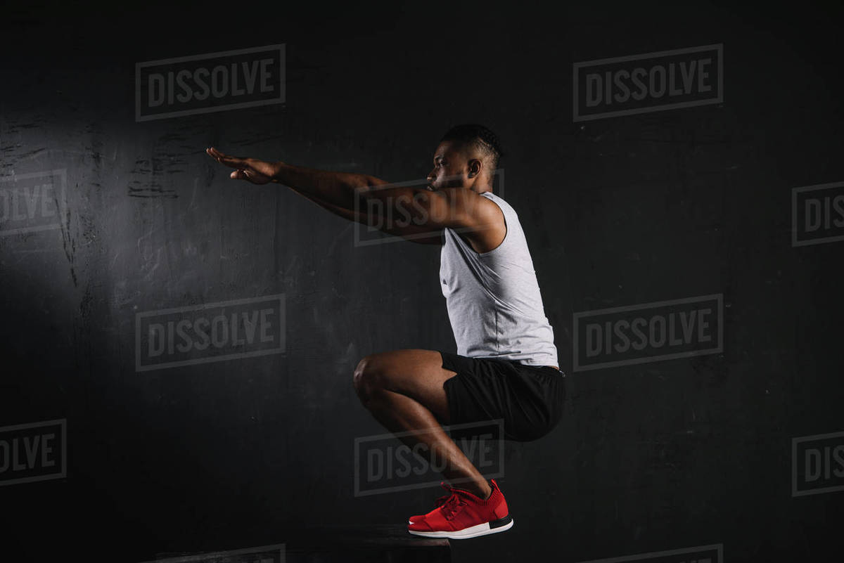 Side view of young African American sportsman squatting on black ...