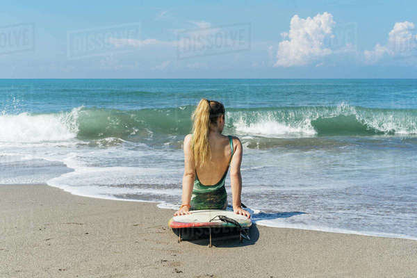 Back view of female surfer in swimsuit sitting on surfboard on beach at ...