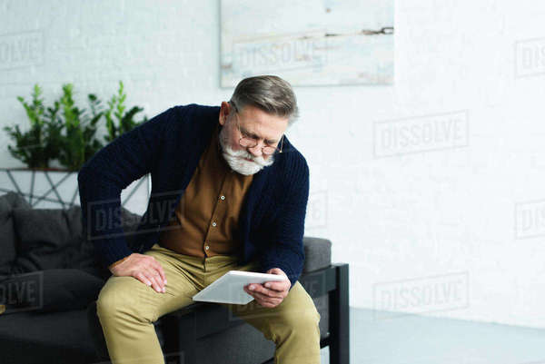 Handsome bearded senior man sitting on sofa and using digital tablet at ...