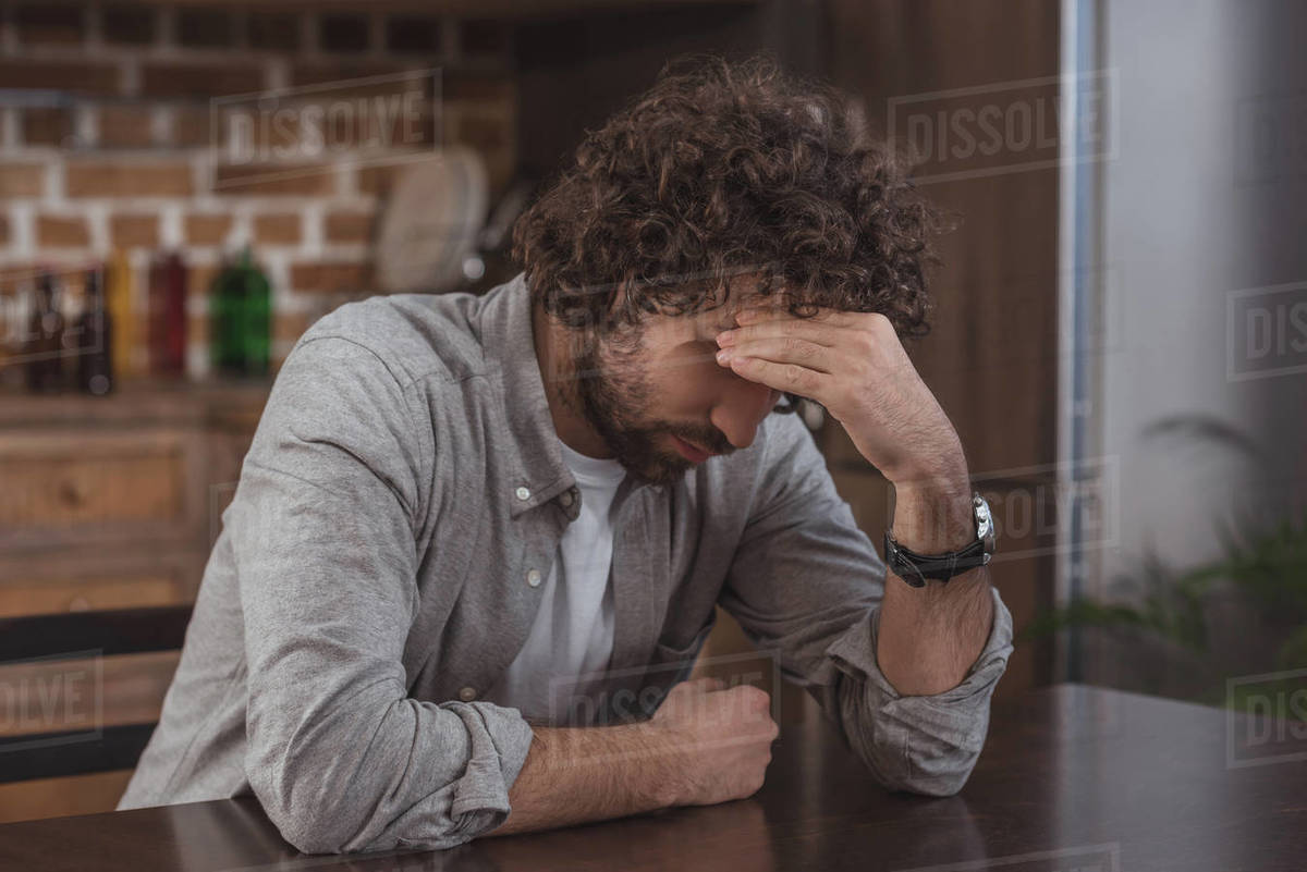 Tired man sitting at table in kitchen and touching head - Stock Photo ...