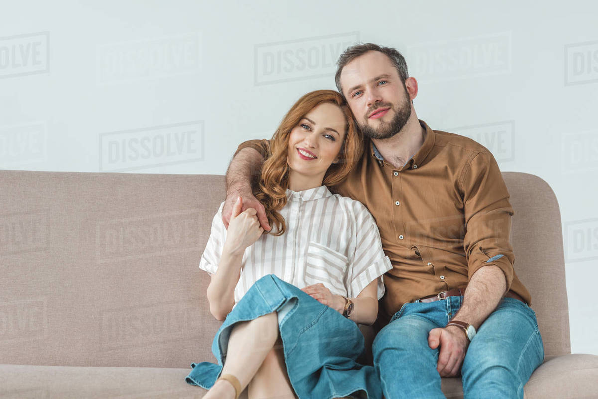 Happy couple sitting on couch and smiling at camera on grey - Stock ...