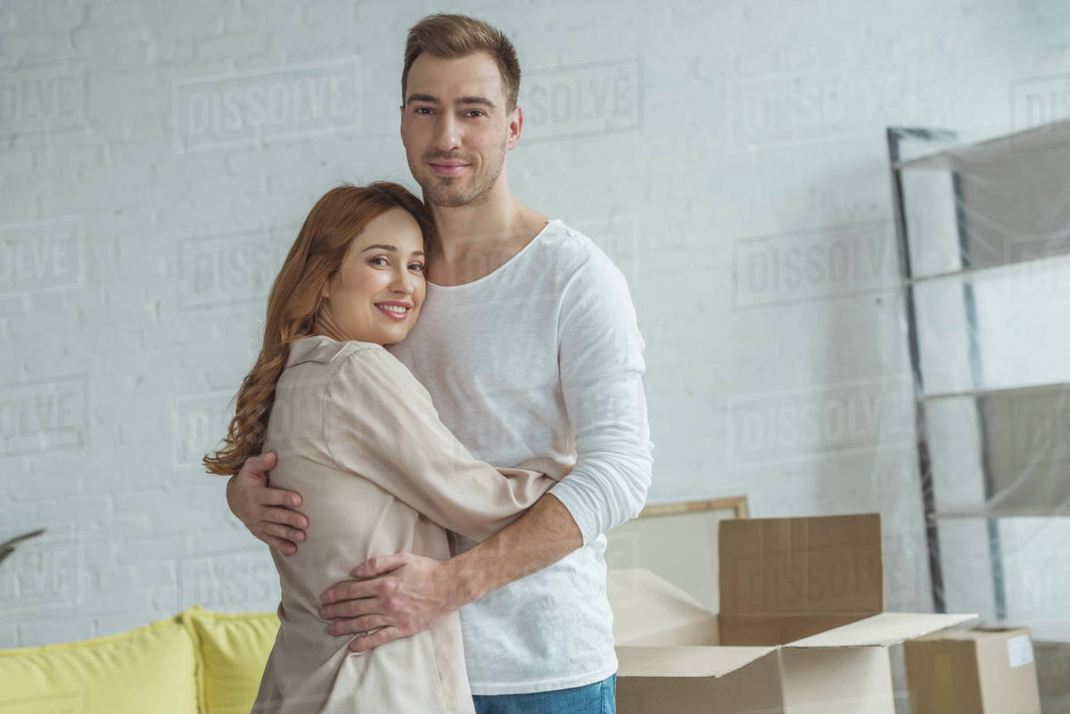 Happy young couple hugging and smiling at camera while moving home ...
