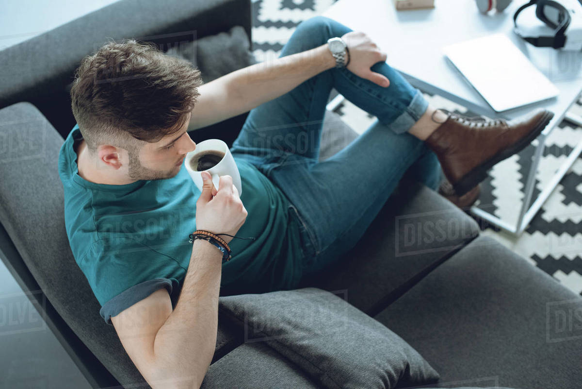 High angle view of young man drinking coffee while sitting on couch ...