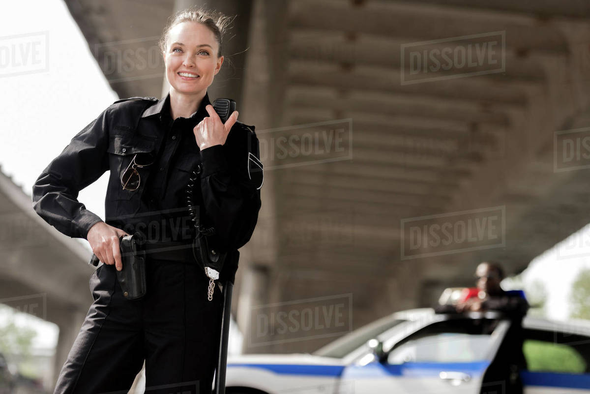 Happy attractive policewoman using walkie-talkie with blurred partner ...