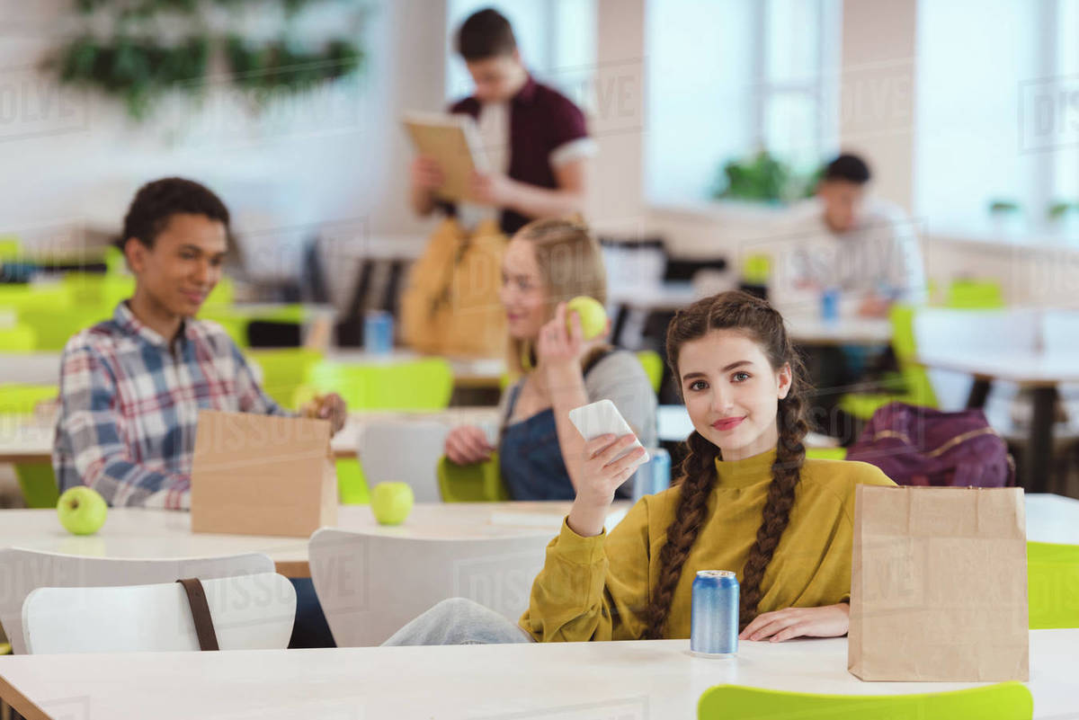 Smiling teen schoolgirl sitting at school cafeteria with classmates and ...