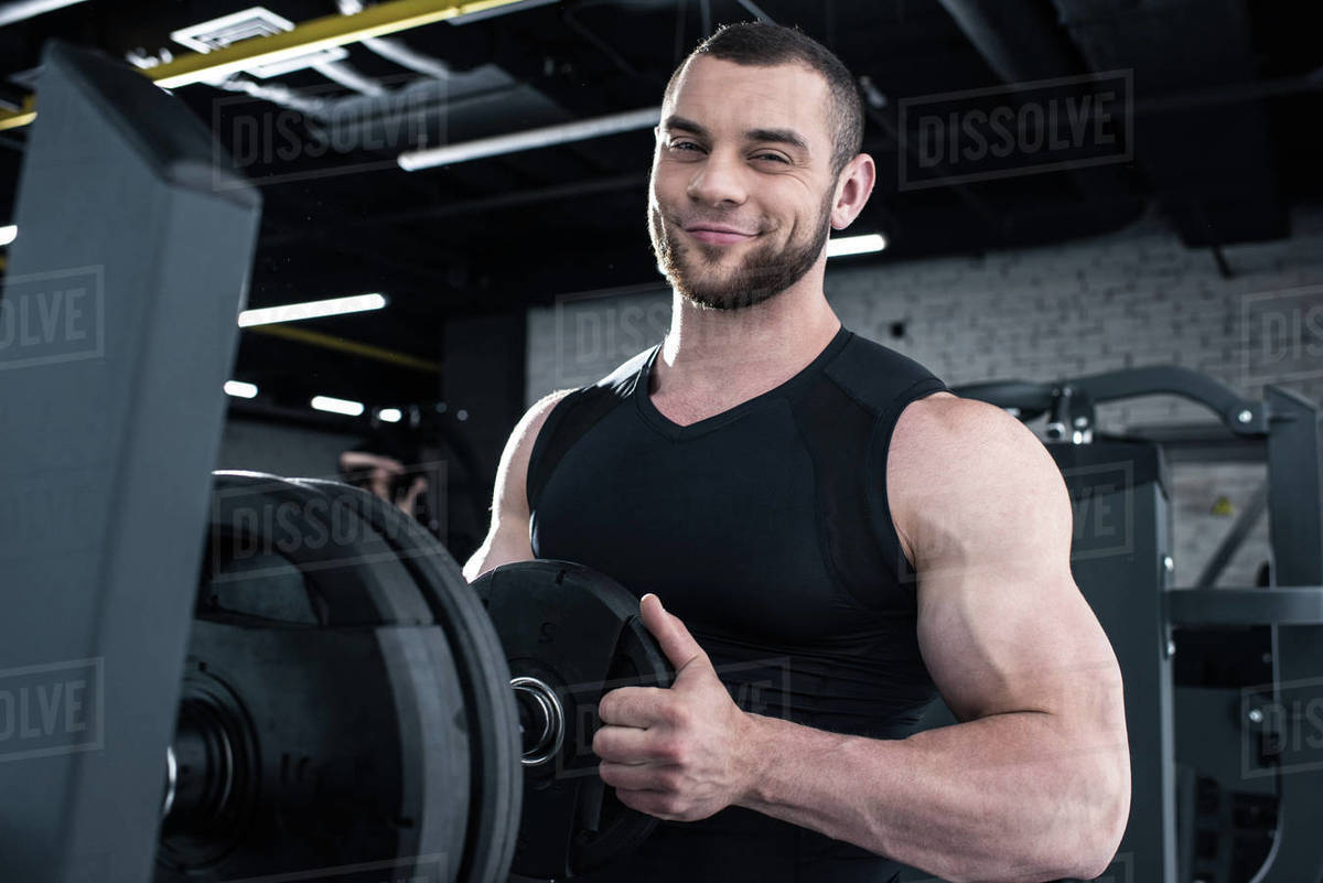 Smiling muscular man putting weight on dumbbell and looking at camera ...