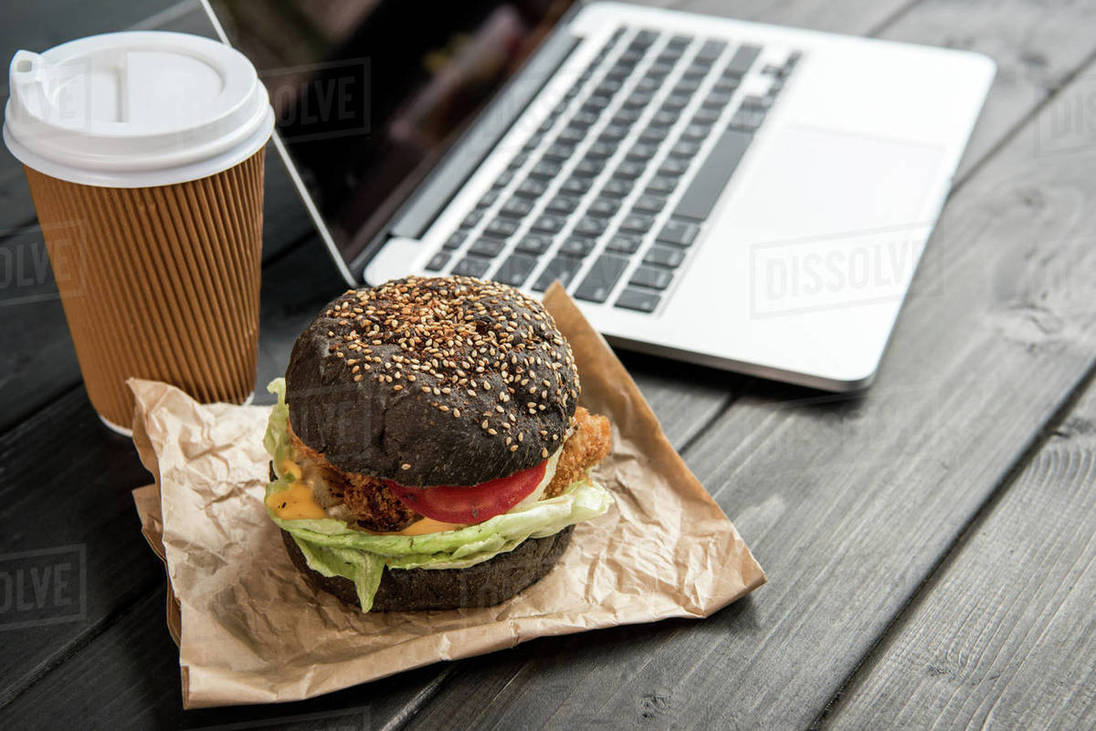 Close up of burger with coffee to go and laptop on wooden tabletop ...