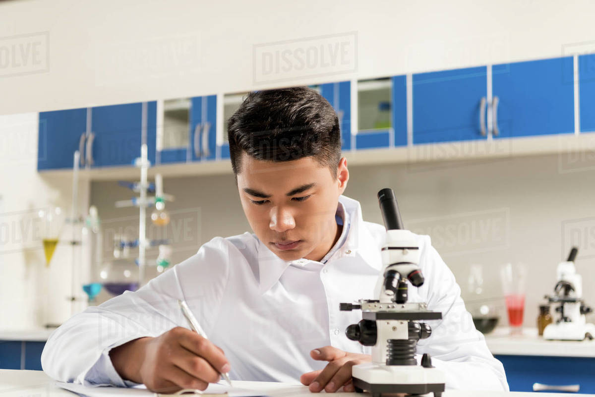 Young lab technician in white coat taking notes on paper after doing ...