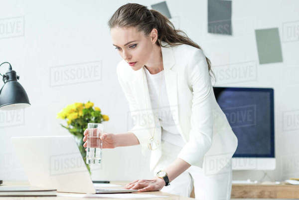 Focused businesswoman with glass of water using laptop at workplace in ...