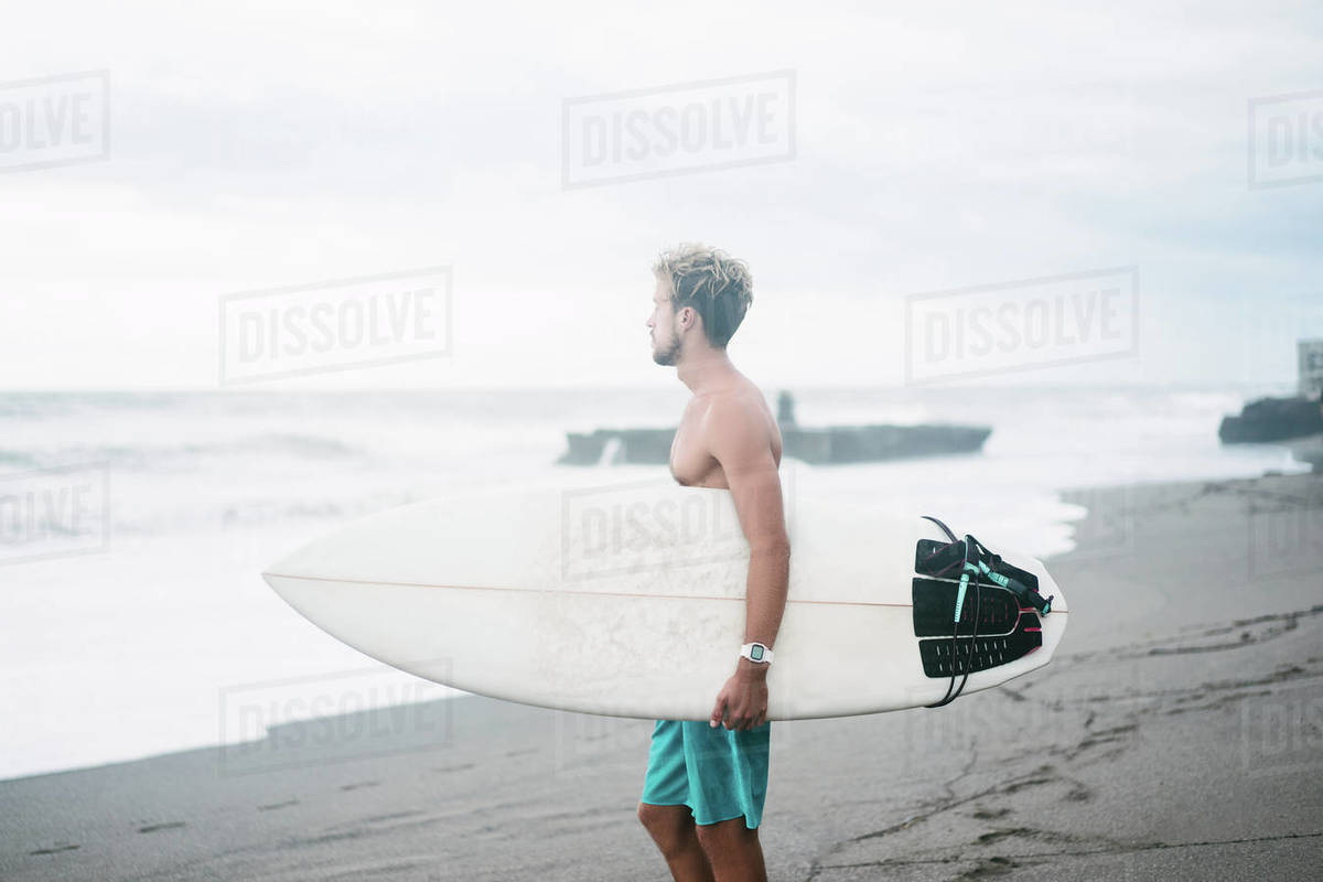 Side view of handsome surfer standing with surfboard in Bali, Indonesia ...