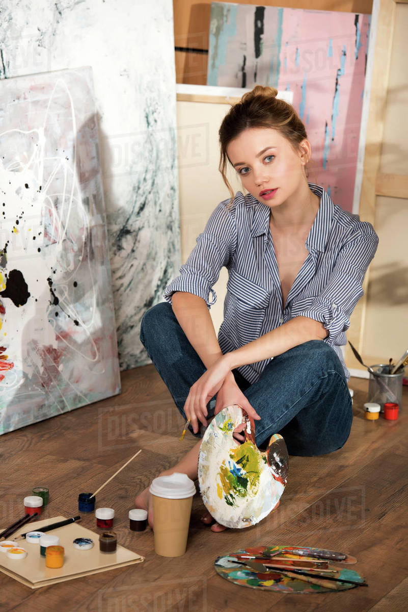 Beautiful young artist sitting on floor and looking at camera in art ...
