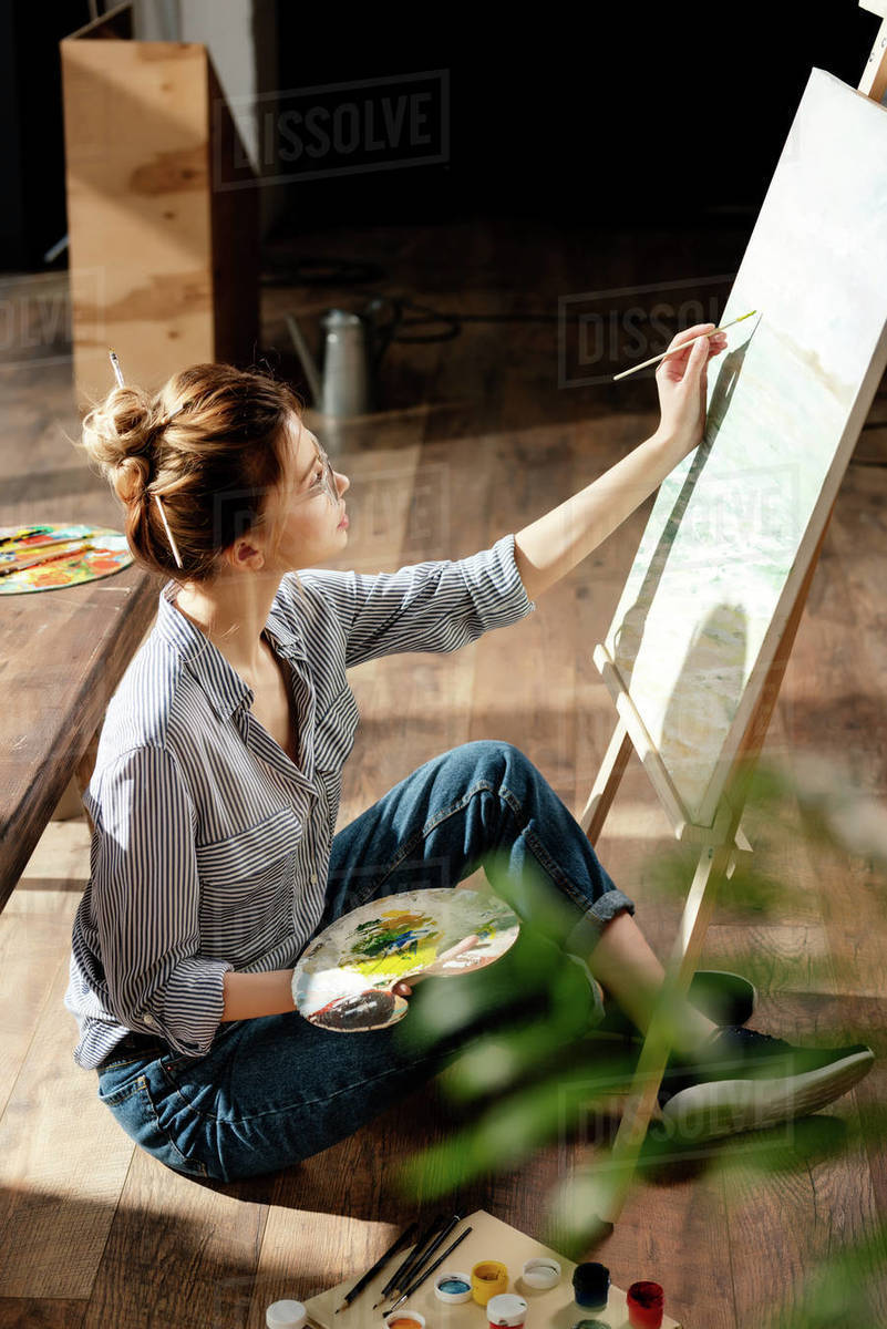 Side view of stylish female artist in eyeglasses painting on easel ...