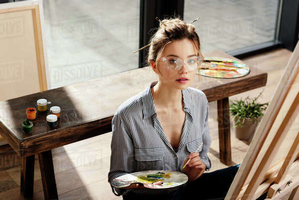 Portrait of stylish female artist in eyeglasses holding palette and ...