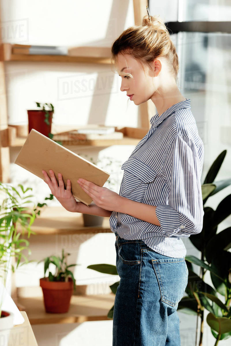 Side view of young woman reading book near potted plants - Royalty-free ...