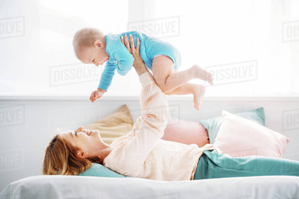 Side view of mother raising laughing little child while lying on bed at ...
