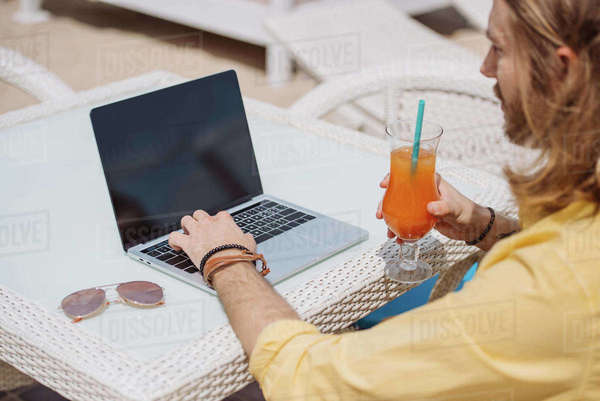 Cropped shot of man holding cocktail and using laptop with blank screen ...