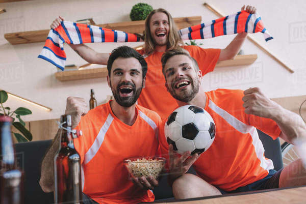 Excited young male football fans in orange t-shirts with scarf, ball ...