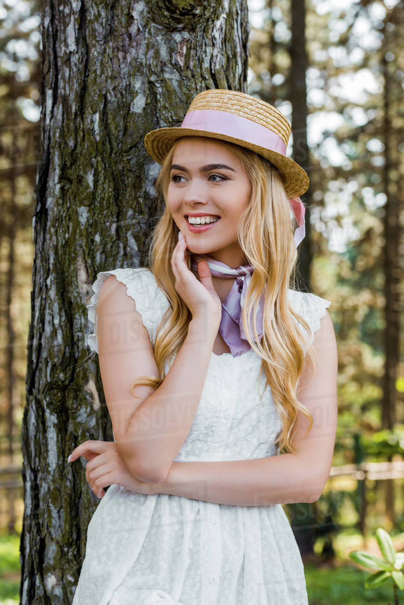 Beautiful blonde girl in wicker hat with ribbon standing near tree and ...