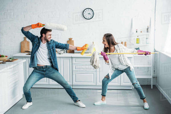 Couple having fun and pretending fight with cleaning tools in kitchen ...