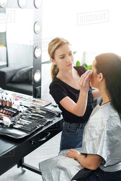 side view of beautiful young woman getting makeup done by focused ...