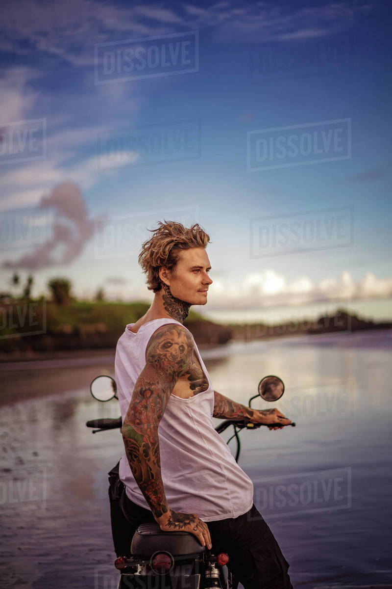 side view of handsome tattooed biker with motorcycle on ocean beach ...