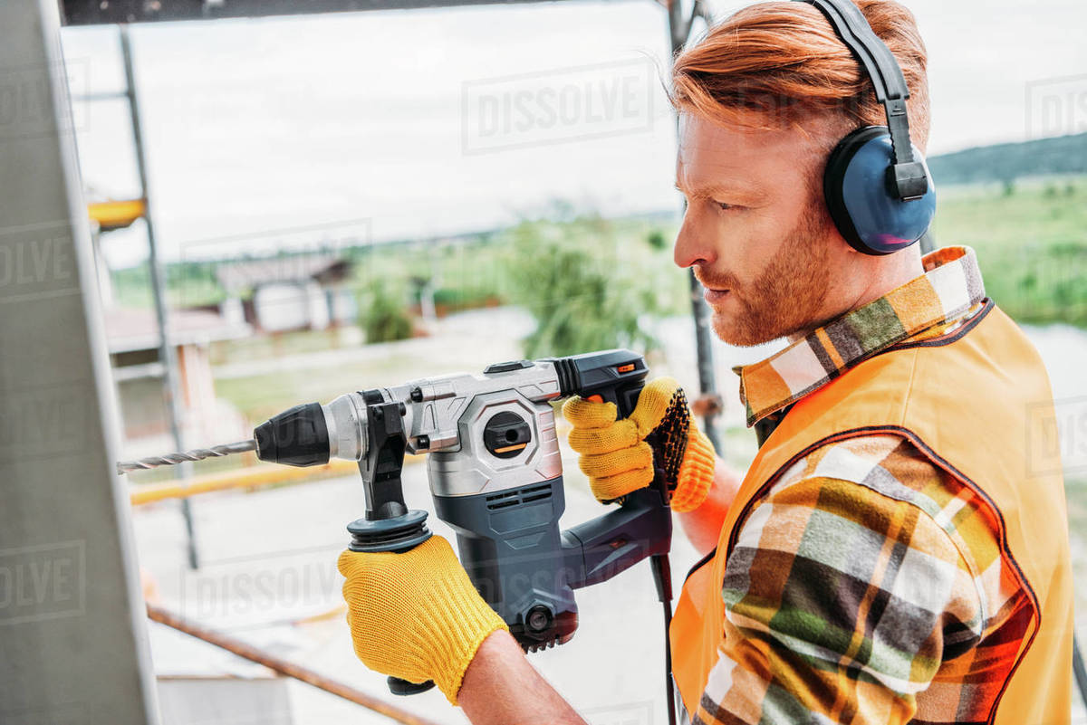 Side view of handsome builder in noise reducing headphones using power ...