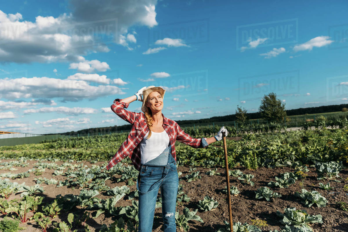 beautiful young female farmer in hat holding hoe and working on field ...