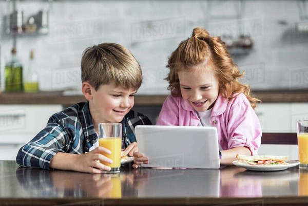 smiling little brother and sister using tablet during breakfast ...