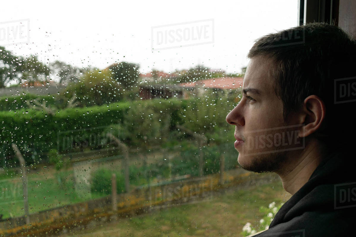 Profile portrait of young man looking out of a window on a rainy day ...