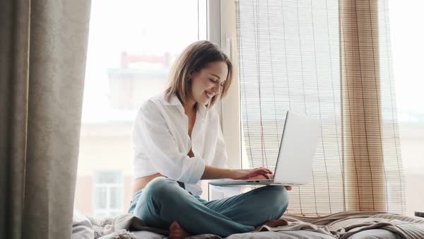 Young woman working on a laptop sitting comfortably on a windowsill at ...