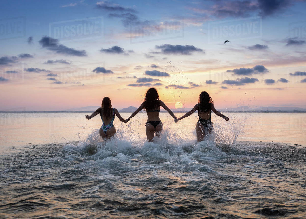 Back view of playful slim girlfriends running in water in bikini at ...