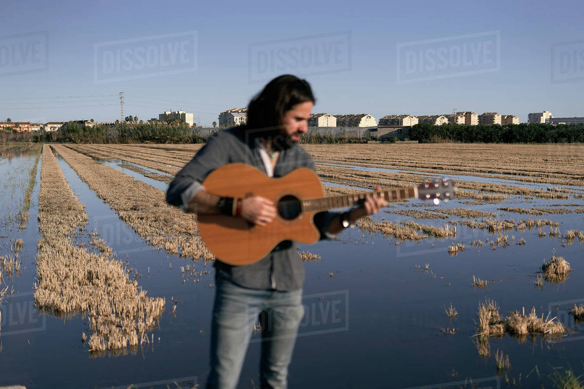 Long haired musician with beard standing on lake shore with guitar and ...