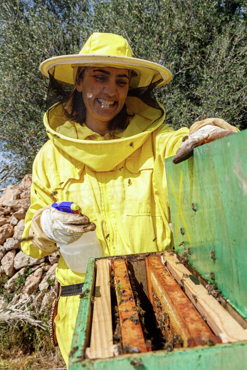 Happy female beekeeper in yellow protective wear using dispenser while ...