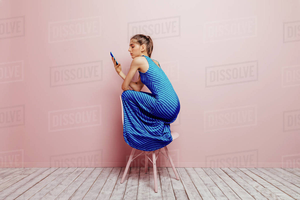 Side view of young woman crouched on top of a chair while using mobile ...