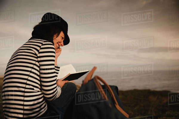 Back view of elegant lady in cap reading volume and sitting on meadow ...