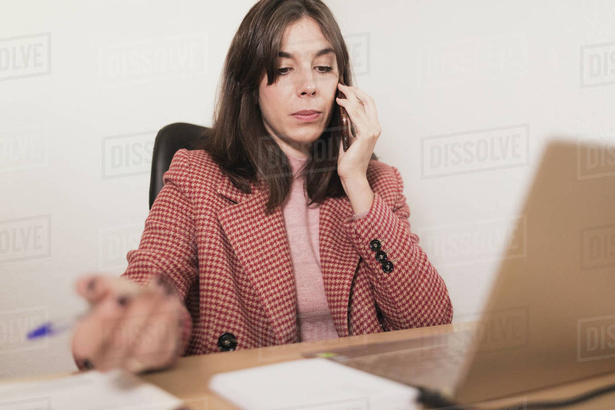 Concentrated young lady using mobile phone and sitting at table with ...