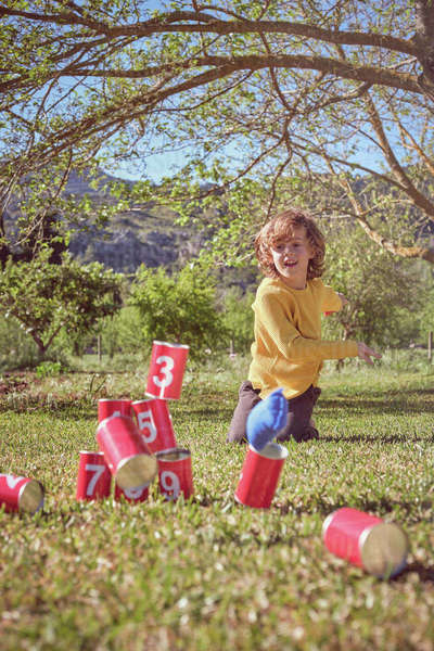 Cheerful kid kneeling and throwing balls in pyramid of red tin cans on ...