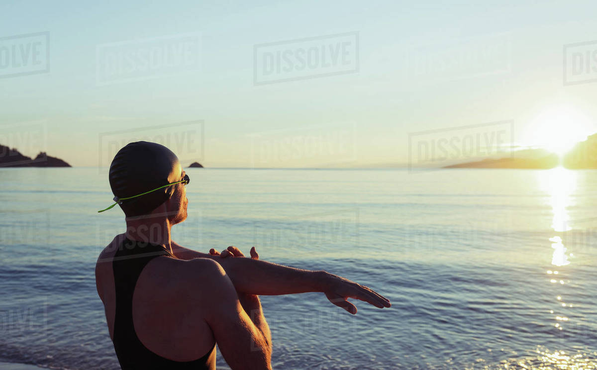 Back view of fit male swimmer in swimsuit and cap standing on seashore ...