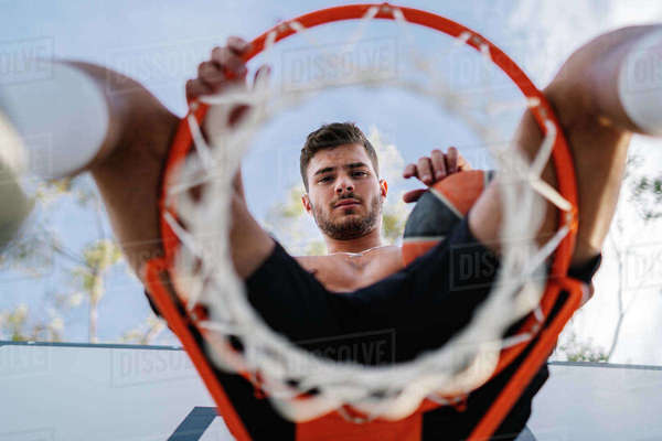 From below of male athlete in sportswear sitting on basketball hoop ...