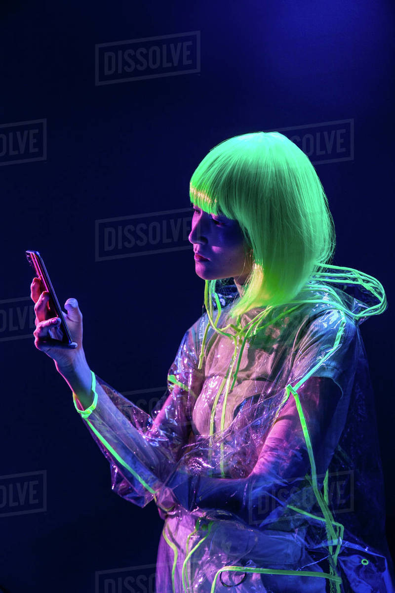 Young asian woman in futuristic wear and green wig using smartphone in ...