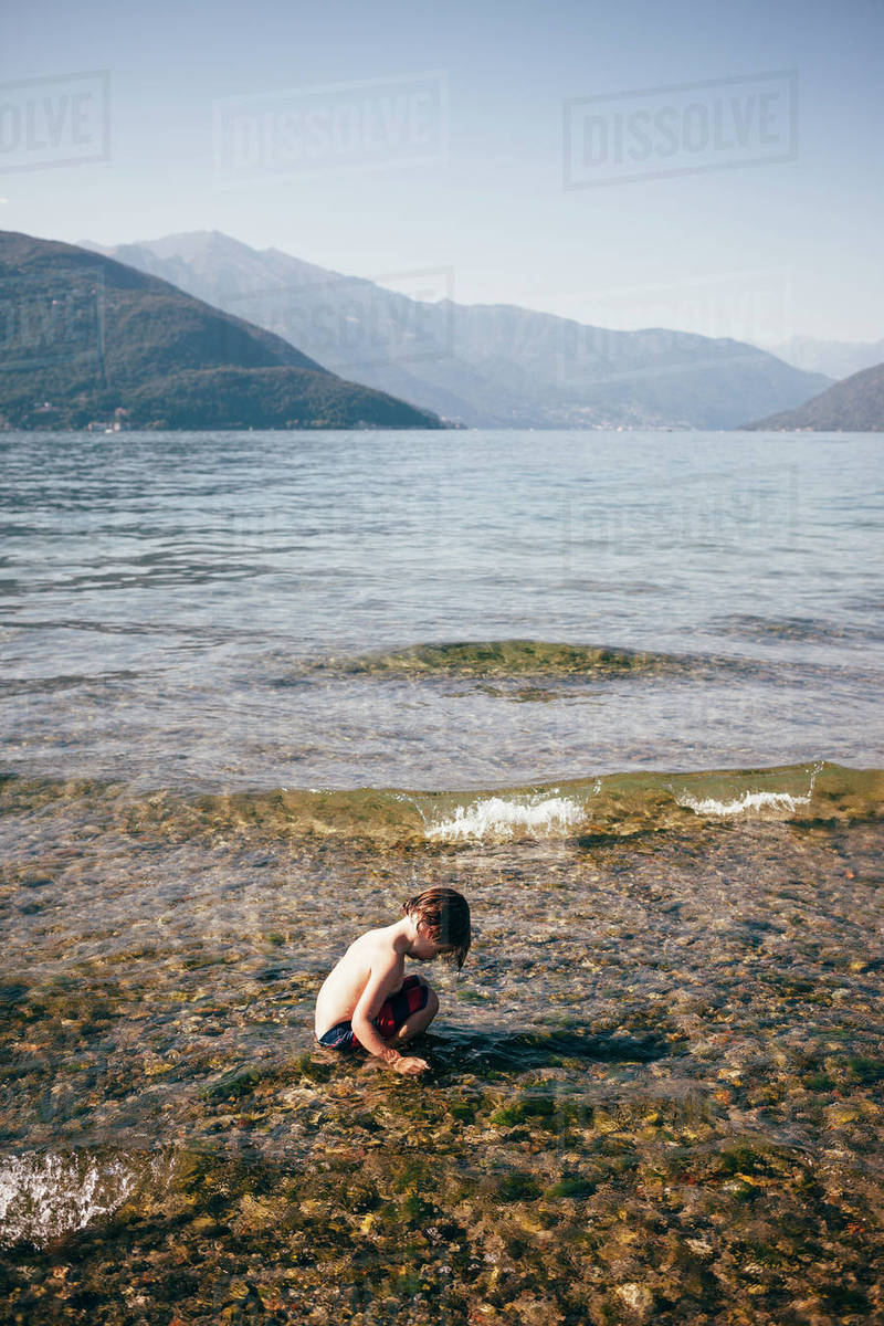 Side view of boy crouching in shallow water looking down and mountain ...